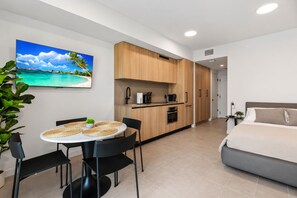 Cozinha moderna com armários de madeira, área de jantar com mesa redonda e cadeiras, e quarto com cama e mesa de cabeceira.