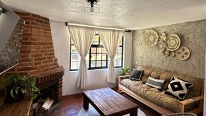 Uma sala de estar com lareira de tijolos, um sofá, uma mesa de centro, cadeiras de vime e um ventilador de teto.