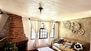 Uma sala de estar aconchegante com lareira de tijolos, mesa de centro de madeira, sofá com almofadas e parede decorada com pratos.