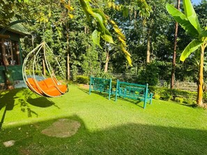 Um balanço e um banco num jardim exuberante com árvores e uma casa ao fundo.