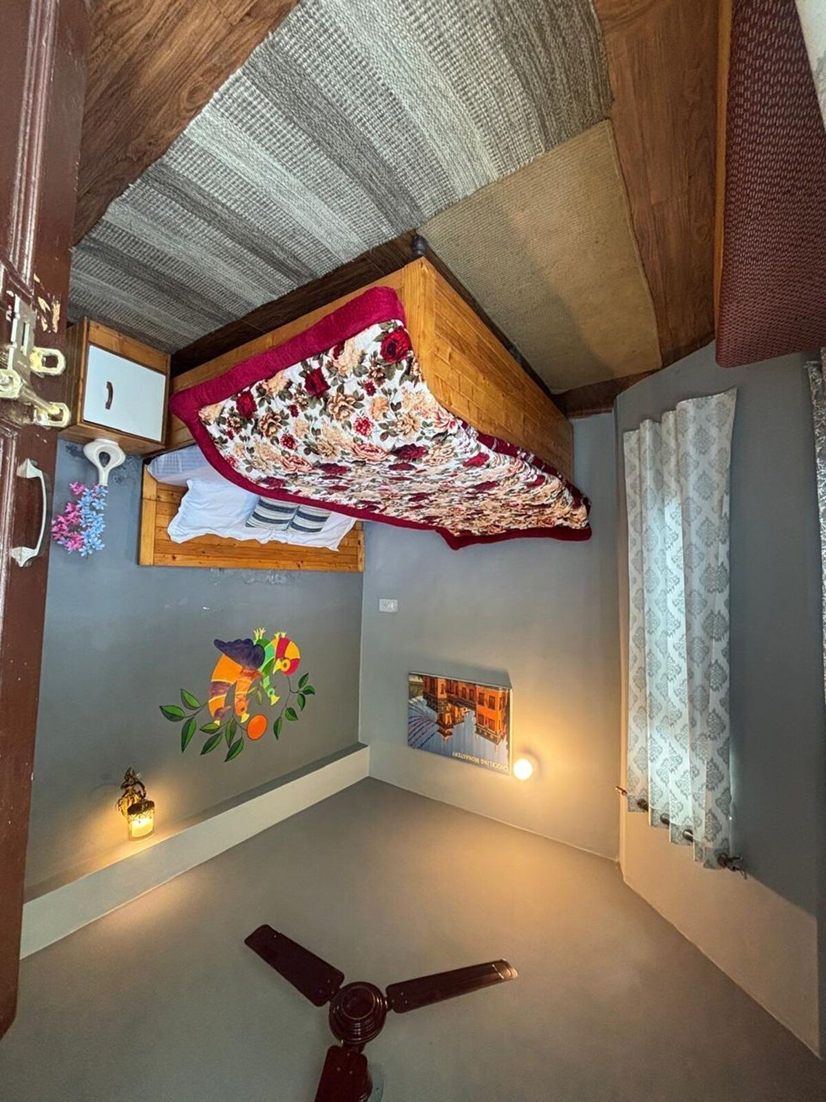Quarto com ventilador de teto, cama com colcha florida, parede com painel floral e quadro emoldurado.