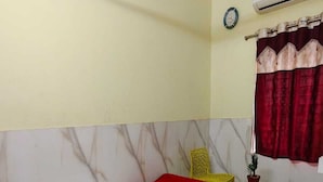 Uma cama de solteiro com estrutura de madeira, um cobertor vermelho e um travesseiro branco em um quarto com piso de mármore e ventilador de teto.