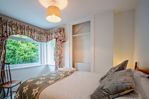 Quarto com cama, cadeira, janela com cortinas, radiador e estante.