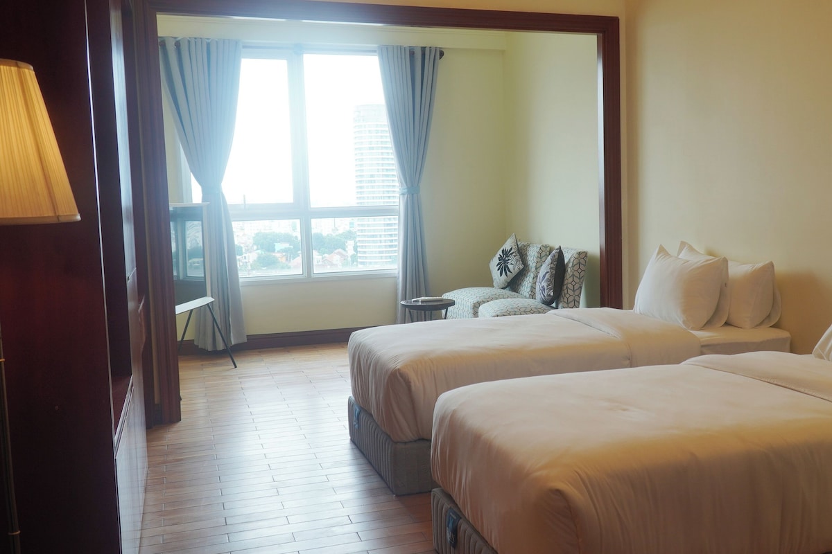 Quarto de hotel com duas camas, uma janela com cortinas e vista para edifícios.