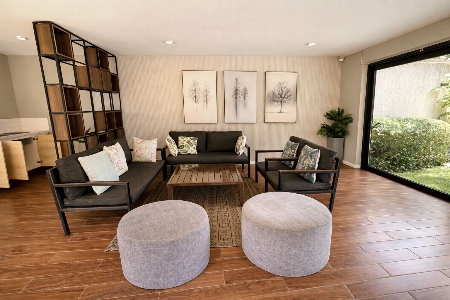 Uma sala de estar moderna com um sofá seccional, duas poltronas, uma mesa de centro e pufes. Há obras de arte emolduradas na parede e uma grande janela com vista para o verde.