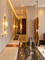 Corredor de hotel com área de estar, luminárias fixadas na parede, uma lixeira e uma porta ao fundo.