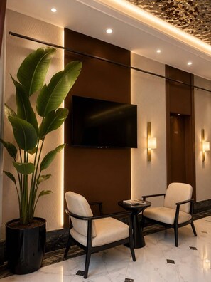 Um hall de hotel moderno com uma grande planta em vaso, duas poltronas, uma mesa pequena e uma televisão de tela plana na parede.