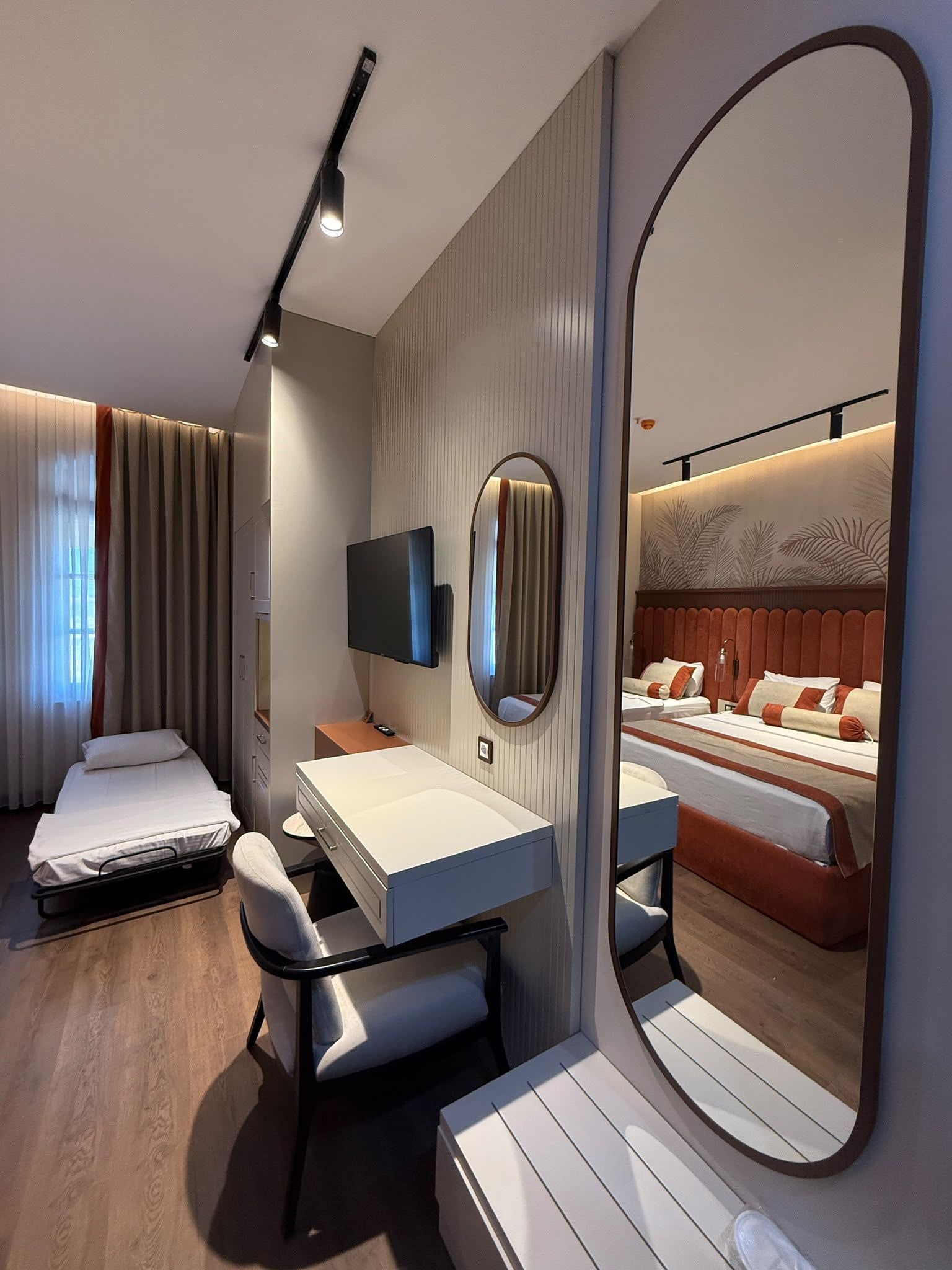 Quarto de hotel com cama, escrivaninha, cadeira, televisão e um espelho grande.