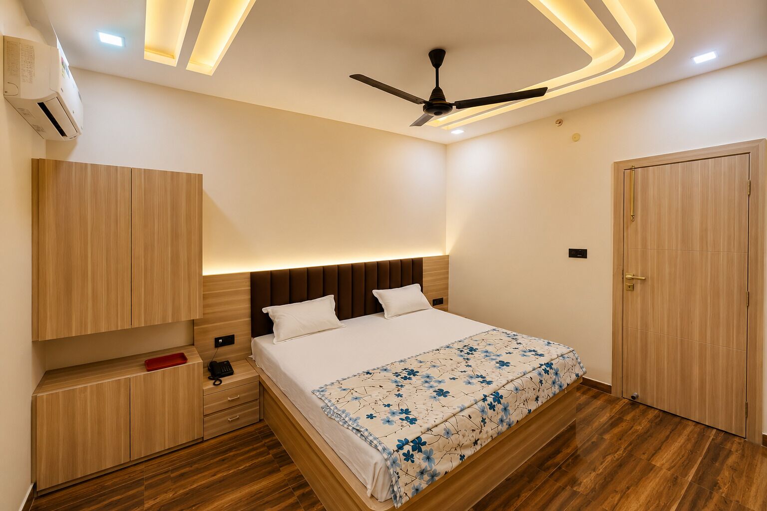 Quarto com cama, mesa de cabeceira, guarda-roupa, ventoinha no teto e porta.