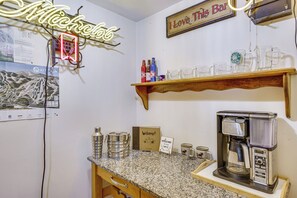 Uma área de bar com um letreiro de neon, prateleiras com garrafas, copos e uma máquina de café.