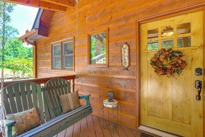 Uma cabana de madeira com varanda, uma cadeira de balanço, uma porta com grinalda e uma pequena mesa com um objeto decorativo.