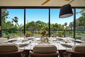 Espaço de refeições com uma mesa posta para seis pessoas, janelas amplas e vista para o verde.