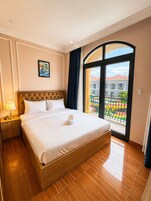 Quarto de hotel com uma cama grande, uma mesa de cabeceira, um quadro na parede e vista para uma área externa colorida através de uma grande janela.