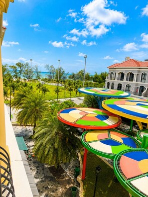Vista de um hotel com áreas de estar ao ar livre coloridas, palmeiras e um edifício com telhado vermelho.
