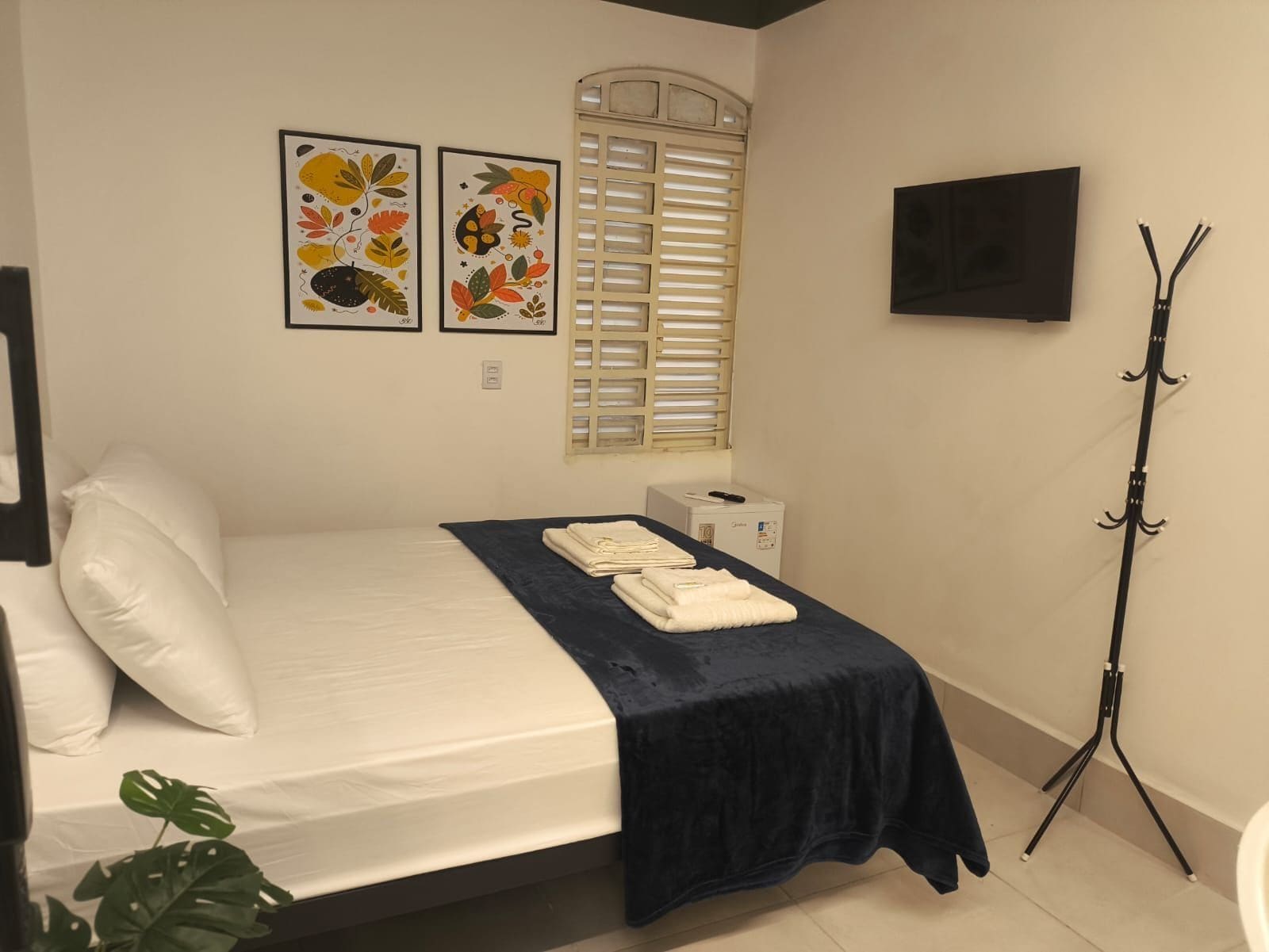 Cama bem arrumada com colcha azul escura e lençóis brancos, uma mesinha de cabeceira com controle remoto, televisão fixada na parede e uma janela com persianas.