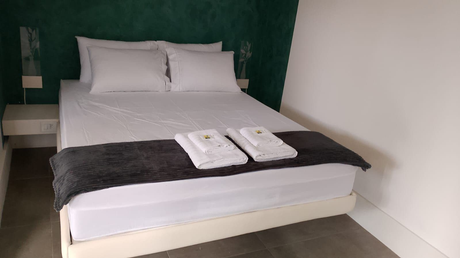 Cama bem arrumada com lençóis brancos e um cobertor cinza escuro, duas toalhas brancas dobradas e uma mesa de cabeceira branca com uma luminária.