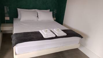Cama bem arrumada com lençóis brancos e um cobertor cinza escuro, duas toalhas brancas dobradas e uma mesa de cabeceira branca com uma luminária.