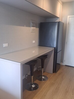 Cozinha moderna com bancada branca, dois bancos altos pretos, frigorífico embutido e uma porta à direita.