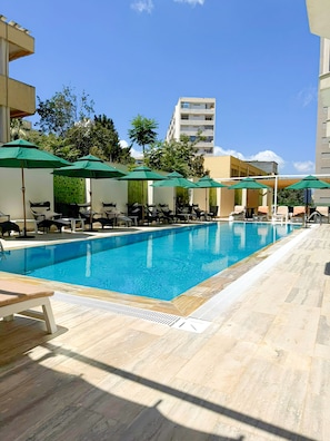 Piscina com espreguiçadeiras e guarda-sóis, ladeada por um deck revestido com azulejos.
