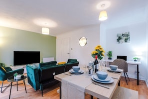 Sala moderna com televisão de tela plana, sofá verde, mesa de jantar para quatro pessoas e pequena cozinha.