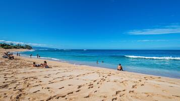 Praia arenosa com pegadas, céu azul límpido e o oceano ao fundo.