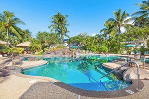 Uma piscina com borda de pedras, espreguiçadeiras e palmeiras.