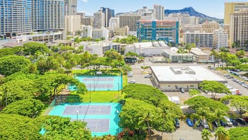 Paisagem urbana com uma quadra de tênis em destaque, cercada por vegetação exuberante e edifícios altos.
