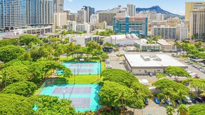 Paisagem urbana com uma quadra de tênis em destaque, cercada por vegetação exuberante e edifícios altos.