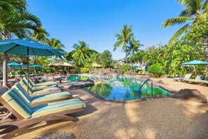 Uma área de piscina com espreguiçadeiras e guarda-sóis, cercada por palmeiras e um céu claro.