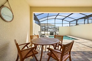 Uma área de pátio com uma mesa redonda de madeira e cadeiras, uma piscina e vista para edifícios ao fundo.