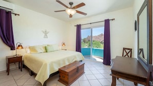 Quarto com cama, luminárias de cabeceira, ventilador de teto, um baú de madeira, escrivaninha e vista para uma piscina e montanhas por uma porta de correr.