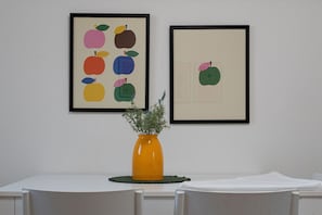 Uma mesa de jantar com cadeiras, uma obra de arte emoldurada com maçãs coloridas e um vaso com folhagem verde.