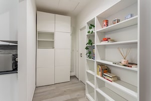 Um corredor com um guarda-roupa branco, uma estante branca com livros e objetos decorativos, e uma porta que dá para outro quarto.