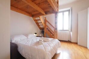 Quarto com teto de madeira, cama com roupa de cama branca, janela com persianas e radiador.