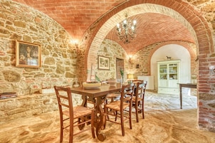 Uma sala de jantar com paredes de pedra, uma mesa e cadeiras de madeira, um candelabro e um armário verde com copos.
