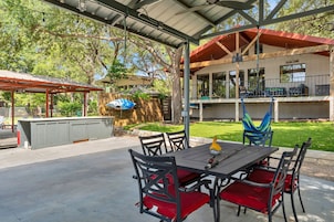 Um pátio externo com mesa e cadeiras, uma área de estar coberta e um edifício com grandes janelas ao fundo.