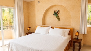 Uma cama grande com lençóis brancos, duas mesas de cabeceira com luminárias, um objeto decorativo na parede e uma varanda com cortinas.