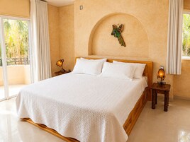 Uma cama grande com lençóis brancos, duas mesas de cabeceira com luminárias, um objeto decorativo na parede e uma varanda com cortinas.