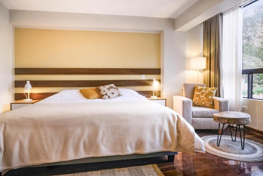 Quarto de hotel com cama grande, mesa de cabeceira com abajur, área de estar com poltrona e mesinha, e vista pela janela.
