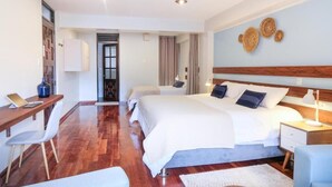 Quarto de hotel com piso de madeira, uma mesa com cadeira, cama com roupa de cama branca e mesa de cabeceira com abajur.