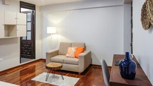 Quarto com sofá, mesa redonda pequena, luminária, cadeira e mesa com um vaso.