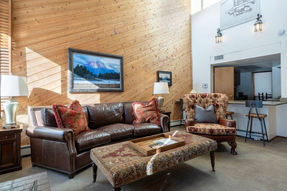 Uma sala de estar aconchegante com parede de madeira, sofá de couro marrom, um banquinho estampado e uma poltrona florida.