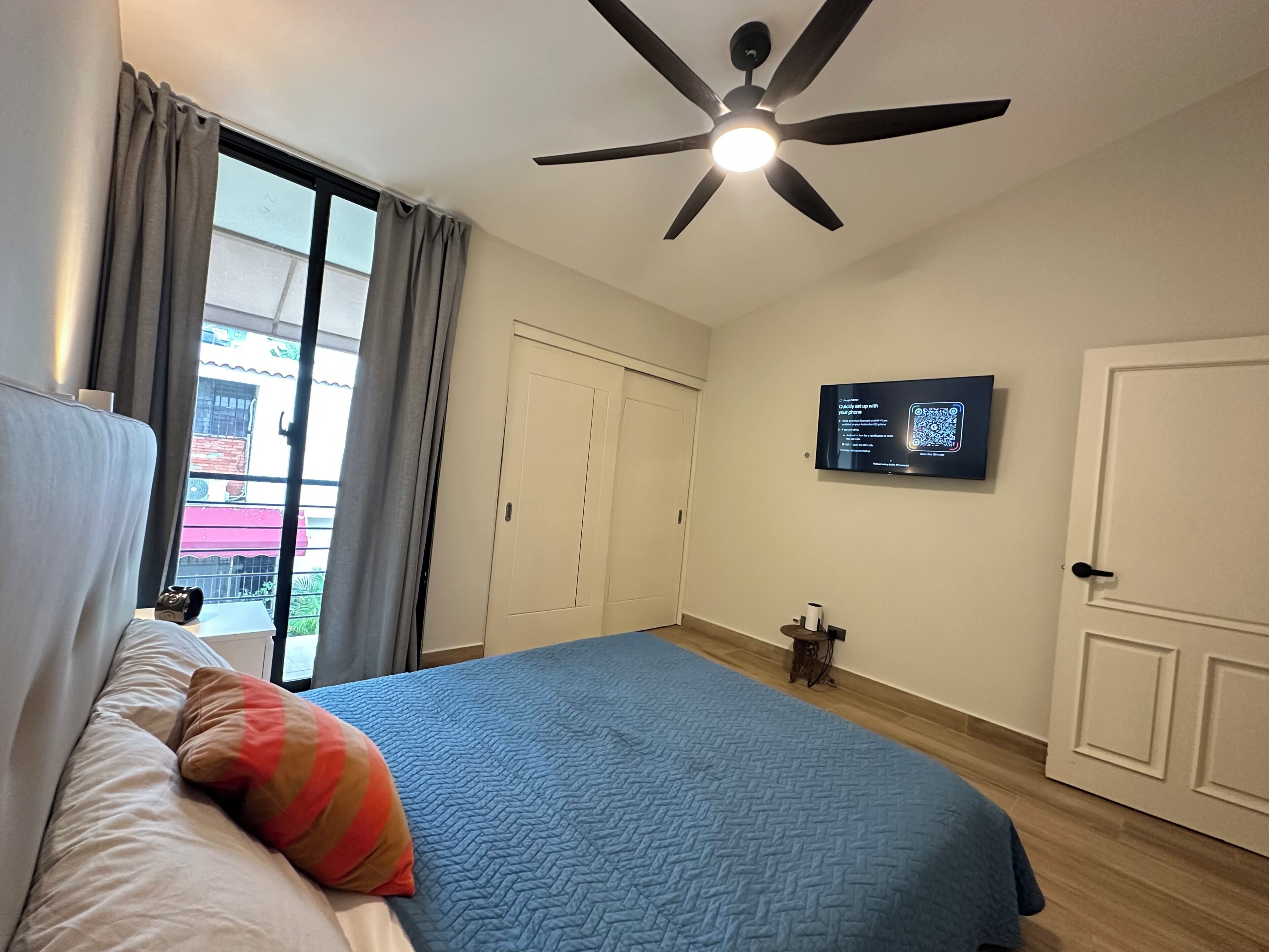 Quarto com cama, ventoinha no teto, televisão na parede e vista para o exterior.