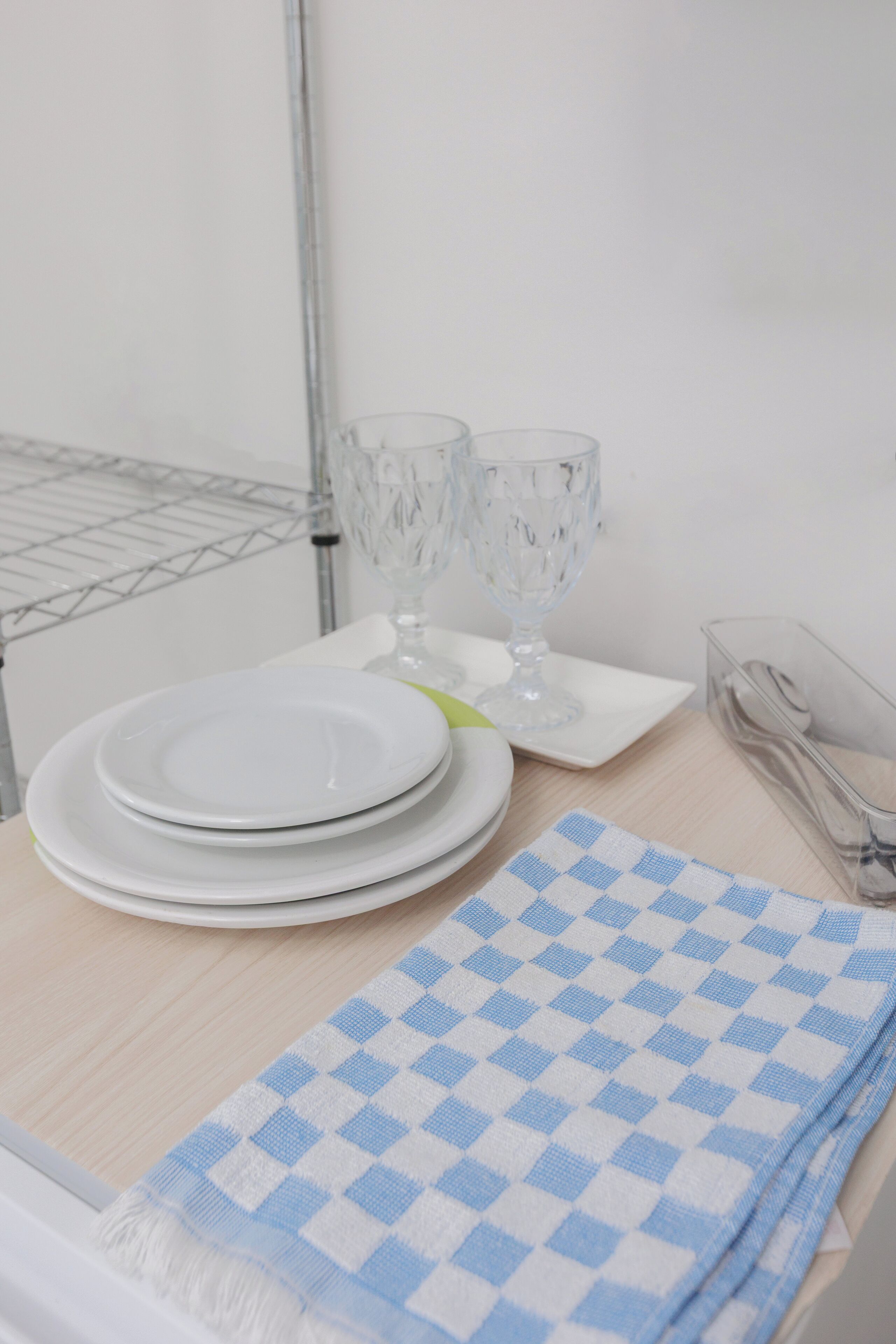 Uma mesa de jantar com pratos brancos empilhados, dois copos de vidro transparentes para vinho, um guardanapo xadrez e uma estante de metal ao fundo.
