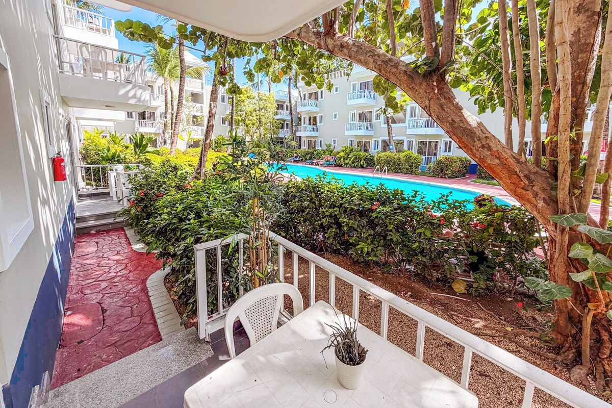 Varanda de hotel com piscina, rodeada por vegetação exuberante e grades brancas.