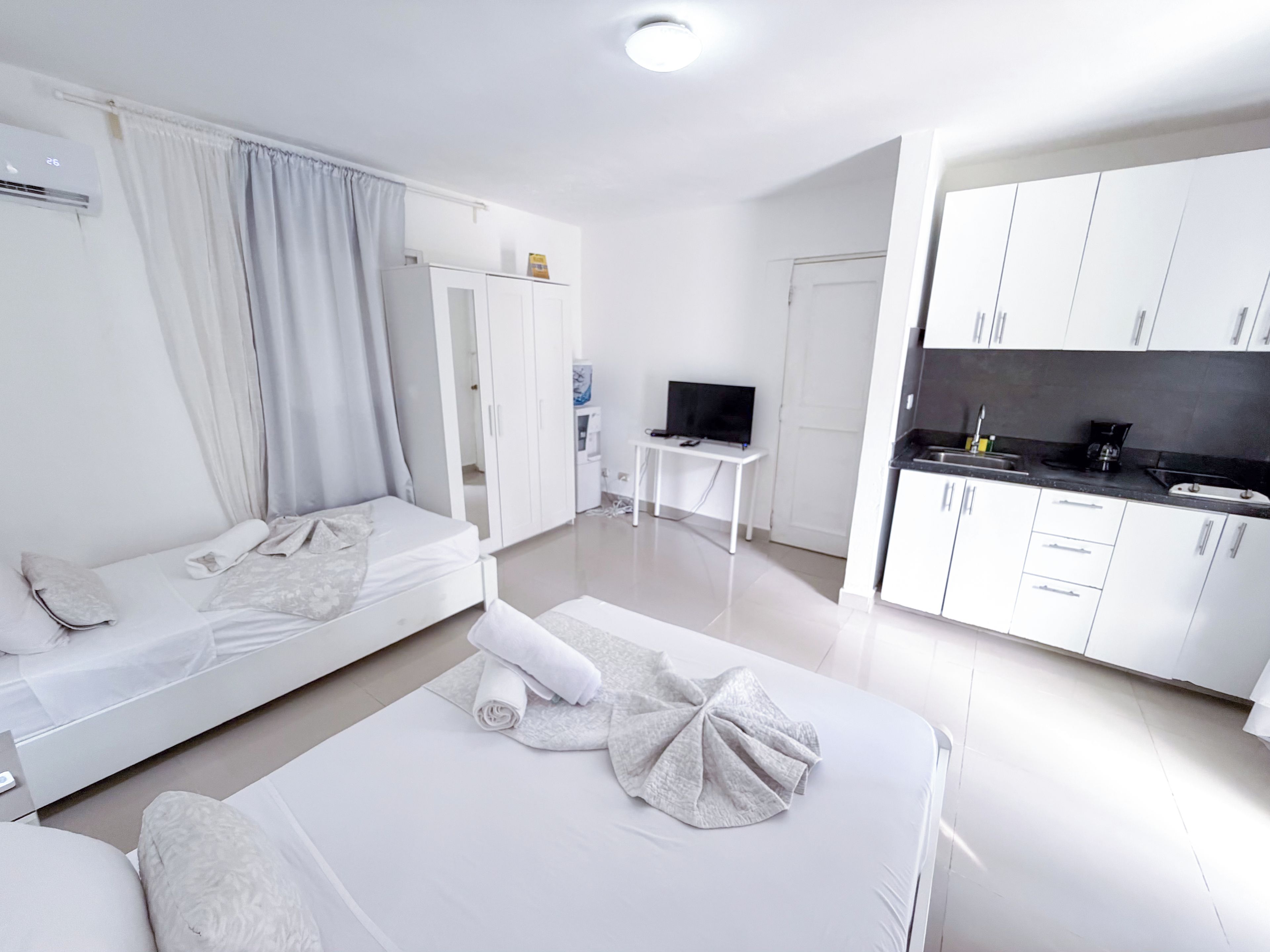 Quarto moderno e bem iluminado, com cama, mesa pequena, televisão e cozinha compacta.