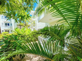 Um jardim exuberante com palmeiras e diversas plantas, um edifício branco com varandas, e um céu azul claro.
