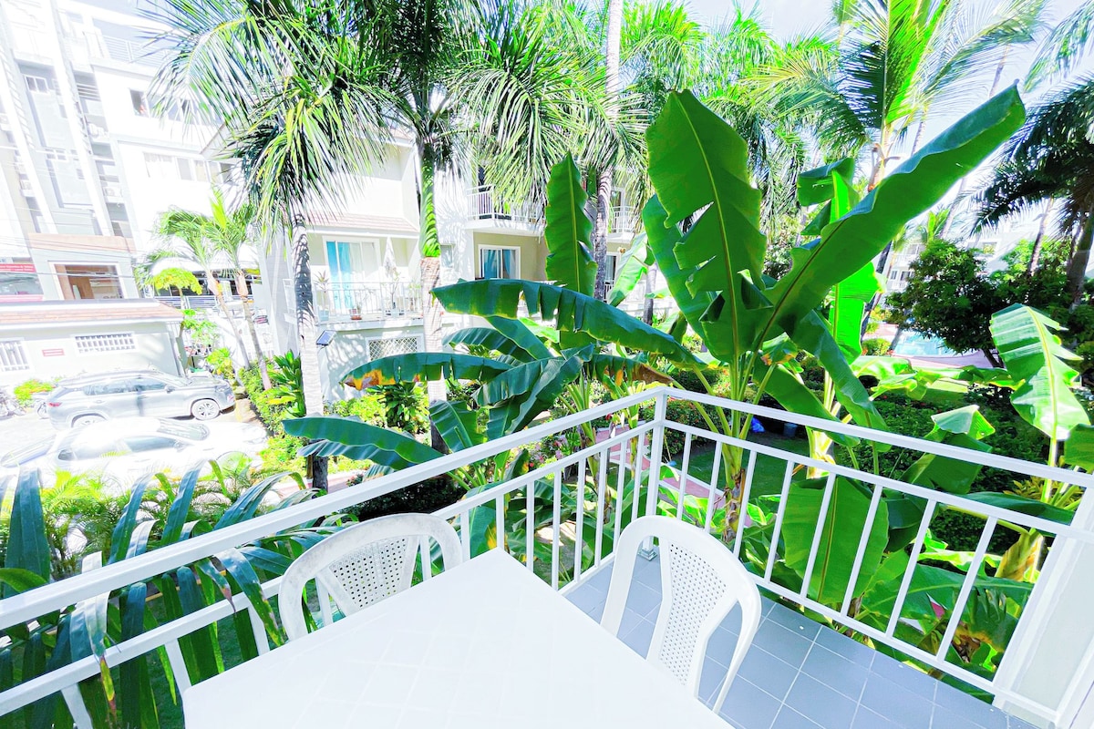 Varanda com cadeiras e mesa brancas, cercada por plantas exuberantes e vista para um bairro residencial.