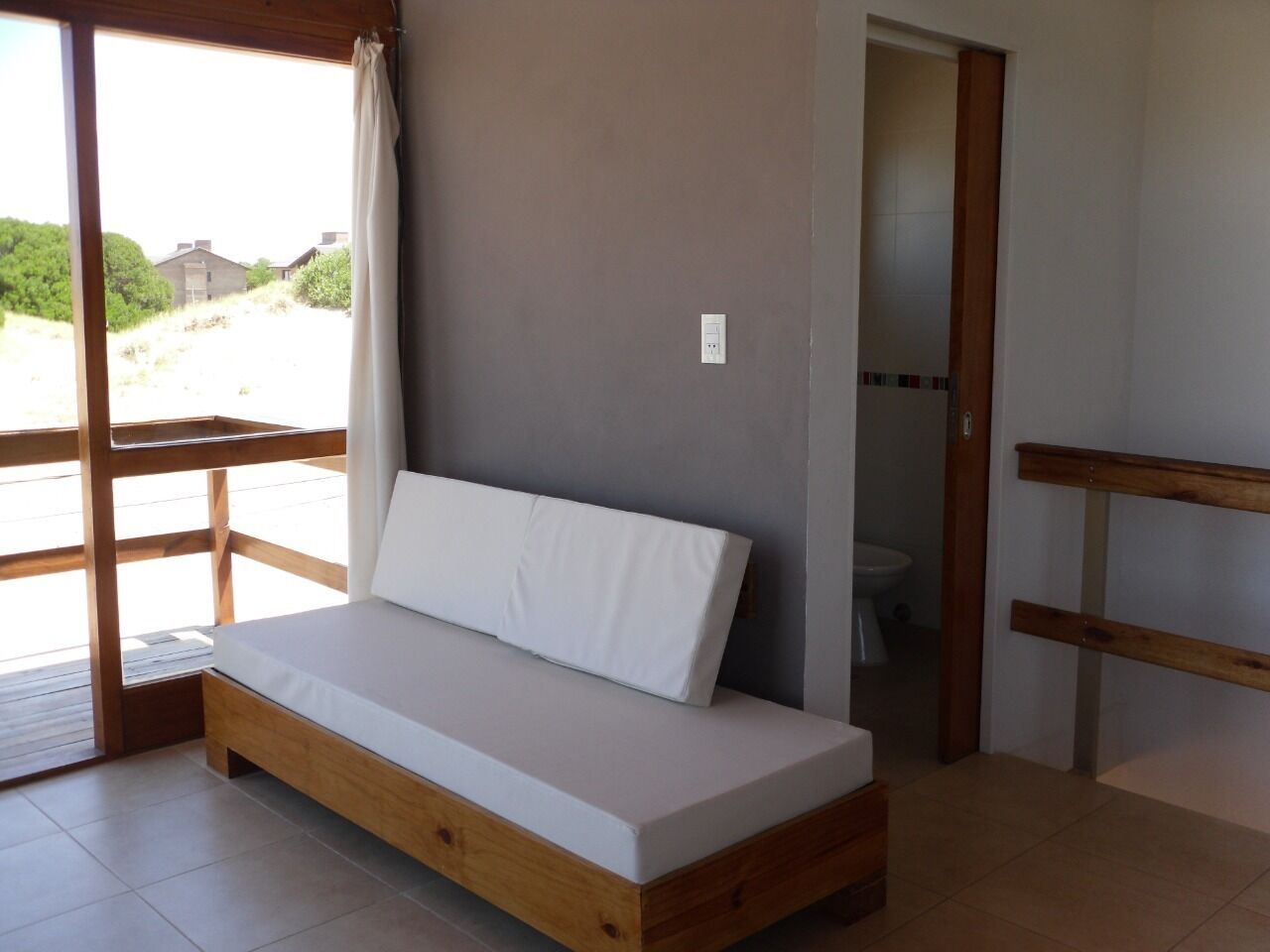 Quarto com cama de madeira, um banco e vista para o exterior.