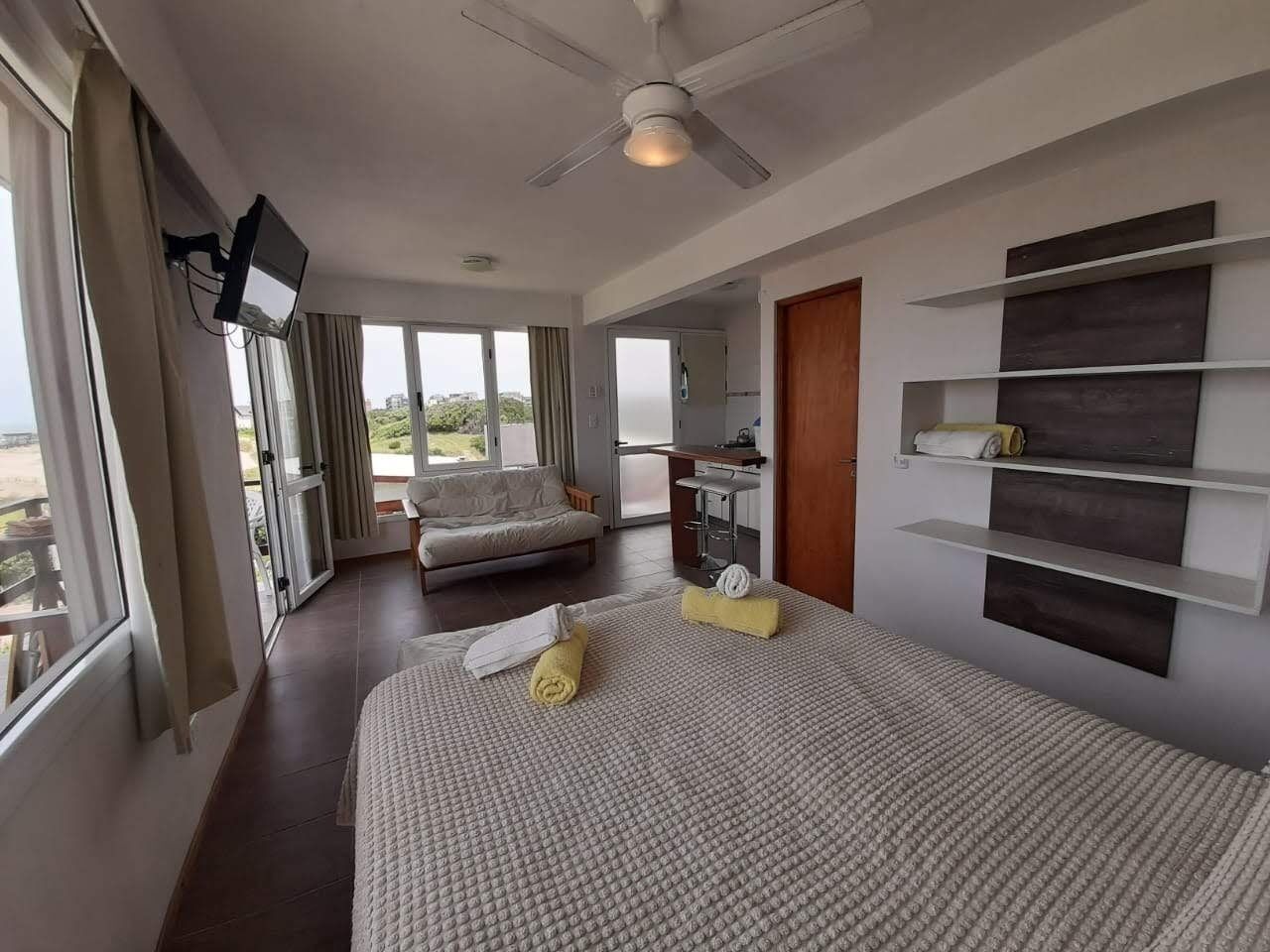 Quarto de hotel com cama, TV fixada na parede, ventilador de teto e vista para o exterior.